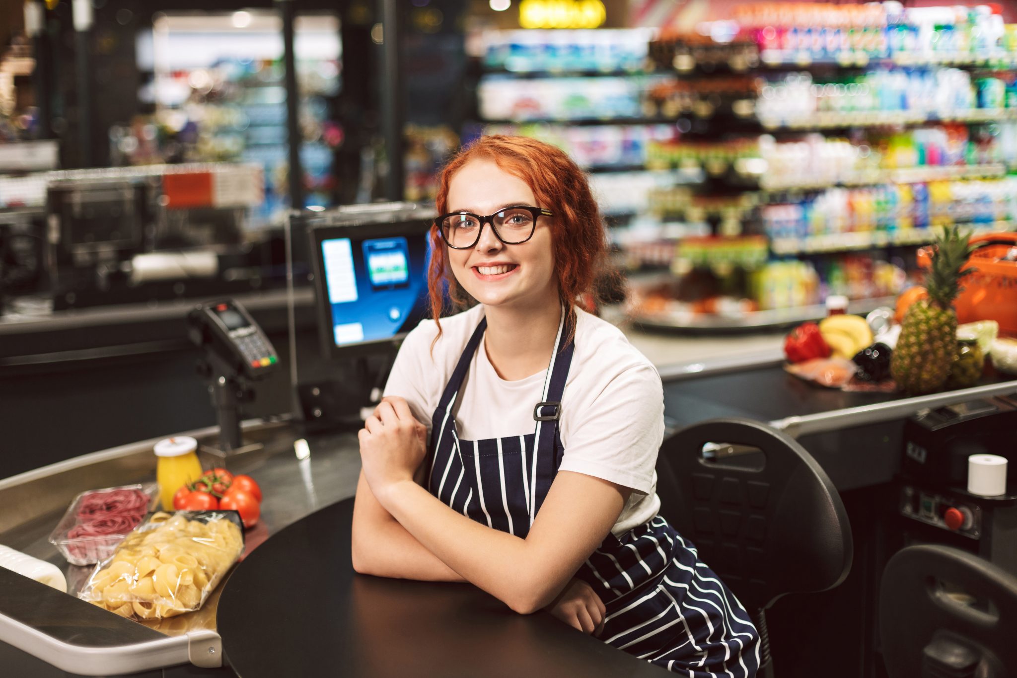 Pretty smiling cashier in eyeglasses and striped apron happily l ...